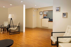 Quarto de hotel com piso de madeira, uma escrivaninha com abajur, duas cadeiras, um espelho e quadros nas paredes.