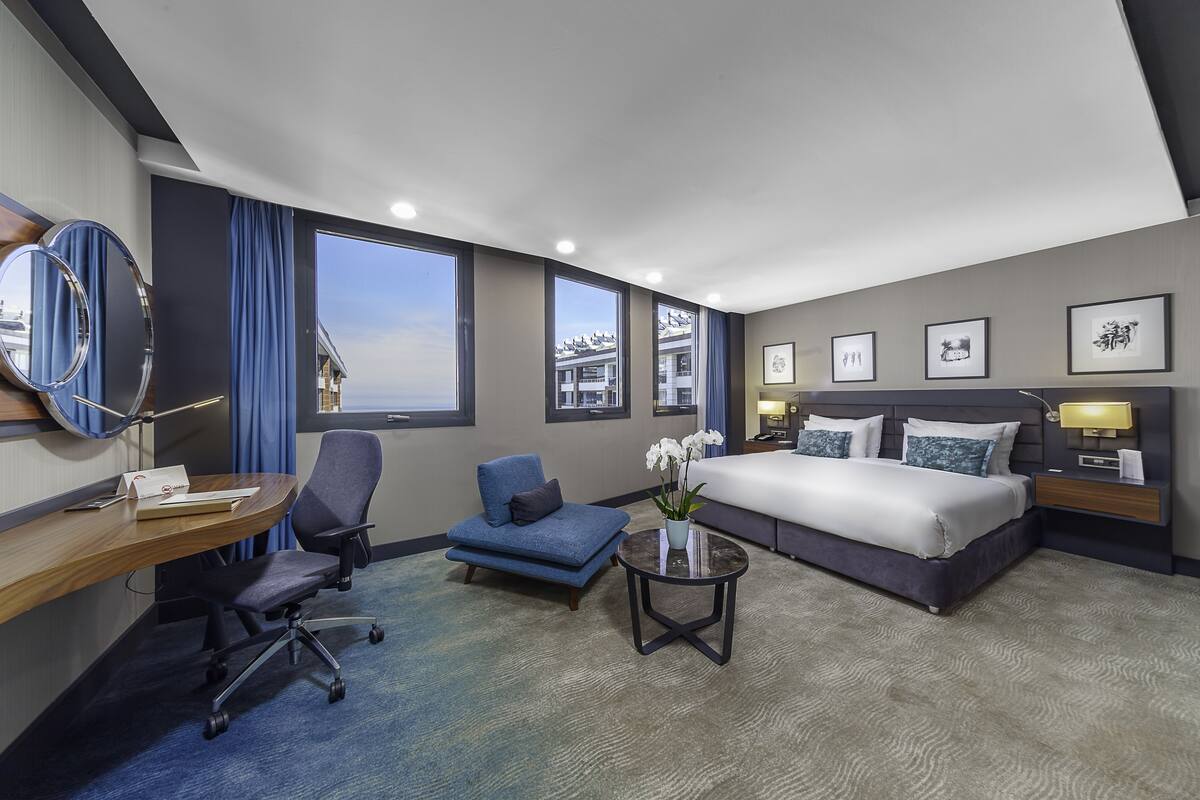Quarto de hotel com uma cama grande, uma escrivaninha com cadeira, uma mesinha com um vaso de flores e vista para edifícios pelas janelas.