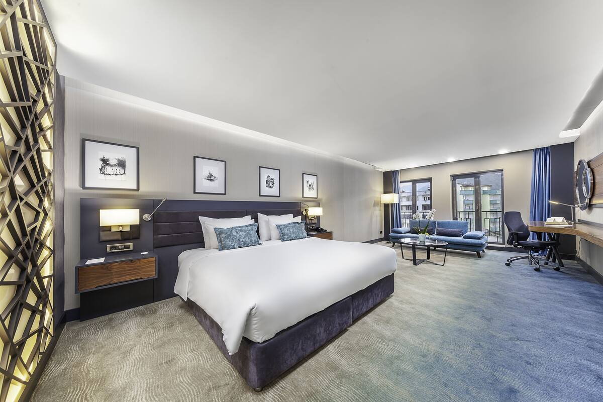 Quarto de hotel moderno com uma cama grande, uma escrivaninha, um sofá e vista para a cidade.