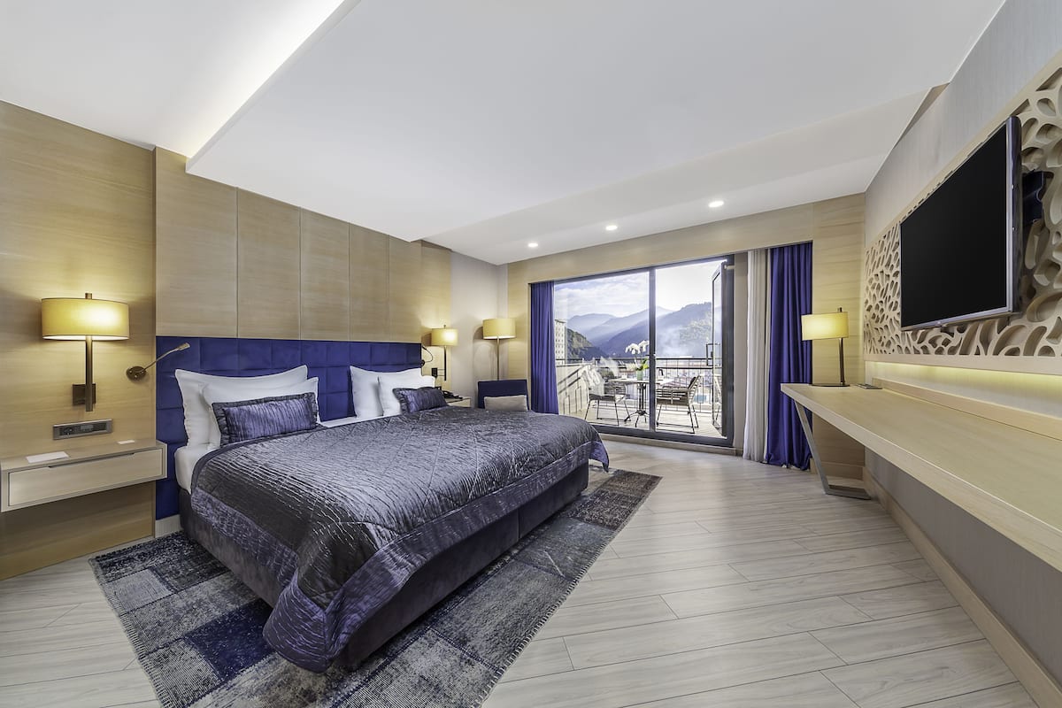 Quarto de hotel moderno com uma cama grande, duas mesas de cabeceira com luminárias, uma televisão de tela plana na parede e uma varanda com vista para as montanhas.