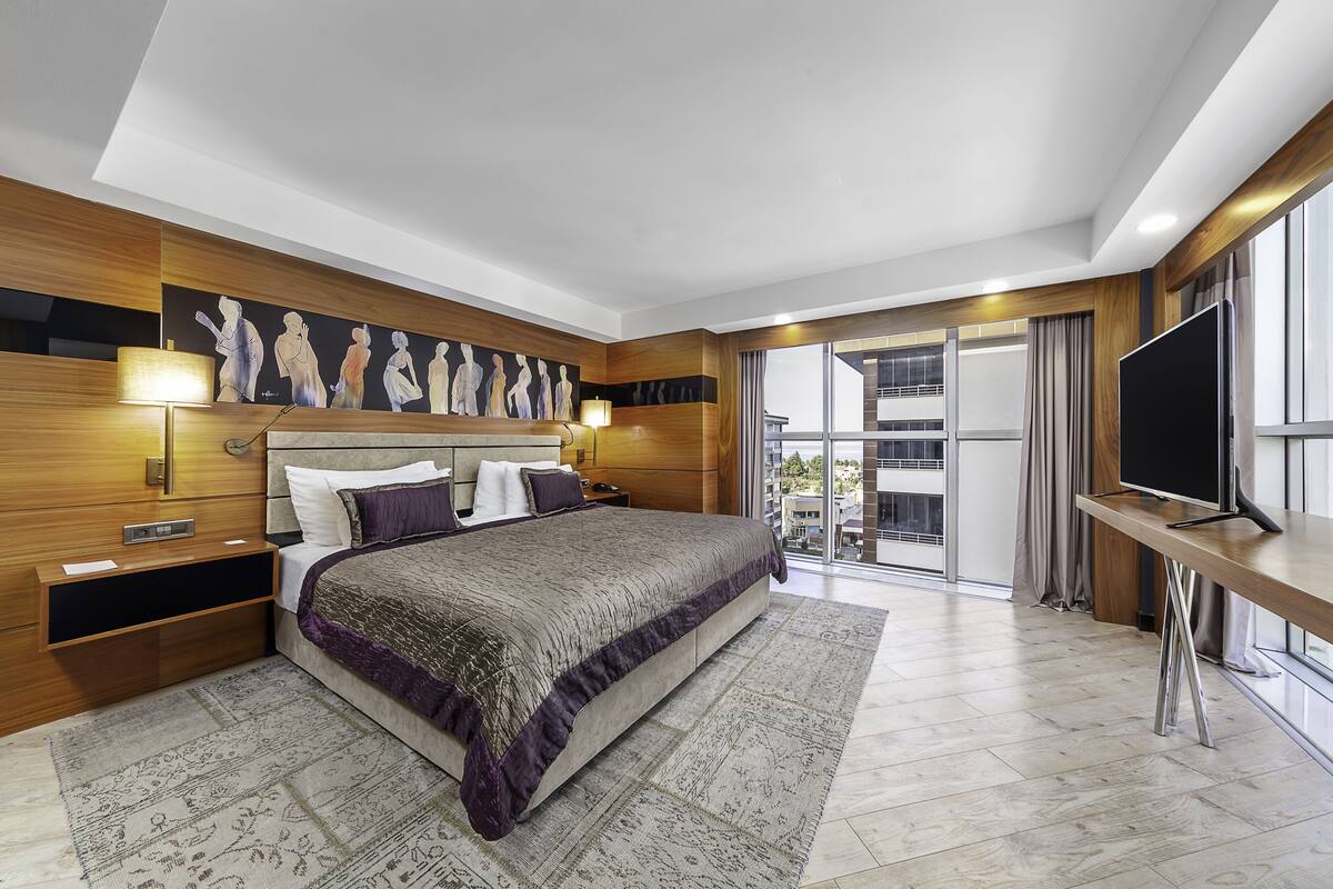 Quarto de hotel moderno com uma cama grande, painéis de madeira, um quadro na parede, uma TV e uma varanda com vista.