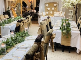 Uma sala de jantar formal com mesas preparadas para refeição, com toalhas de mesa brancas, talheres dourados e decoração com folhagens verdes.