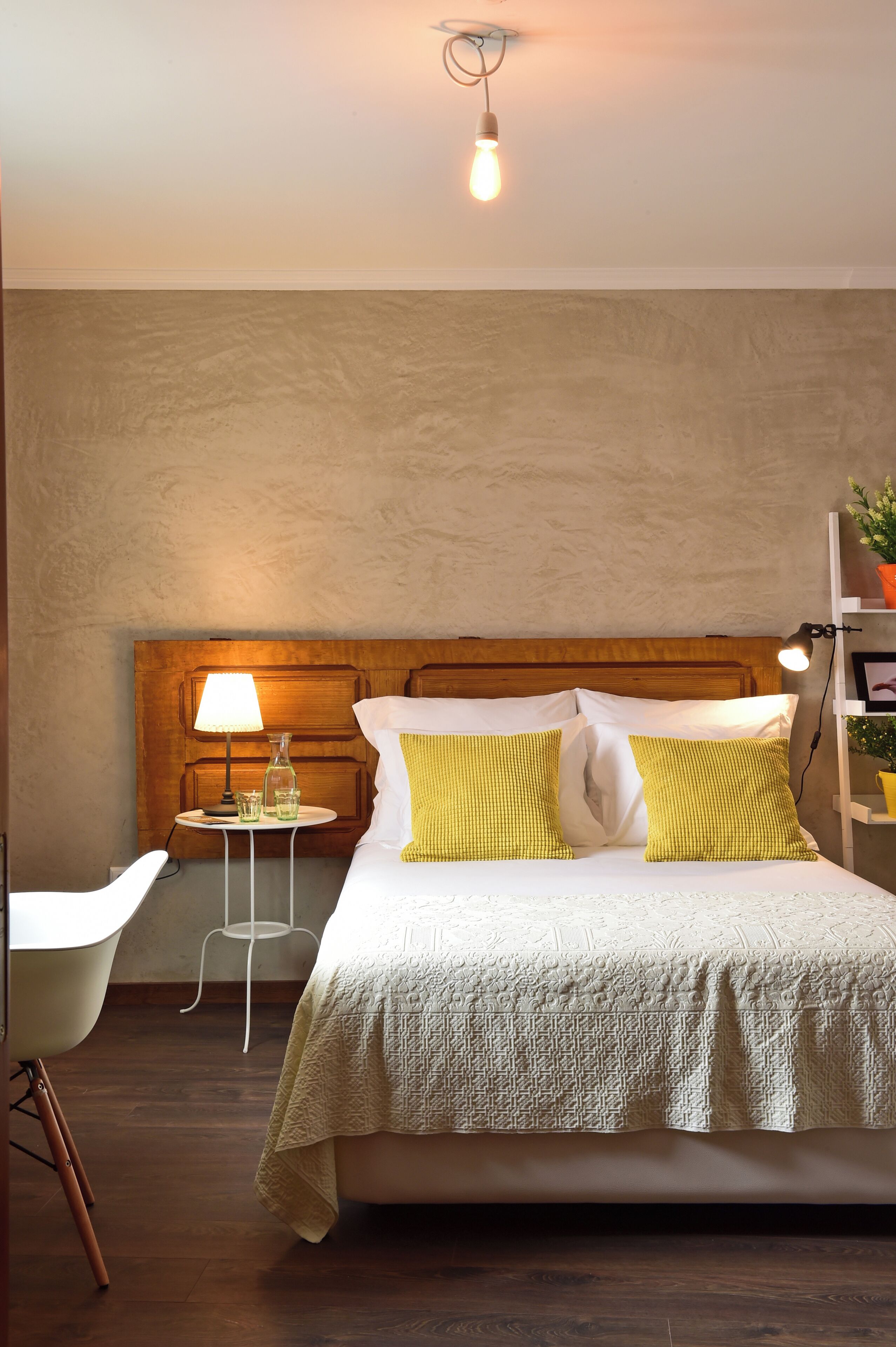 Quarto com cabeceira de madeira, cama com roupa de cama branca e amarela, mesa de cabeceira com abajur e uma pequena mesinha com abajur e uma garrafa.