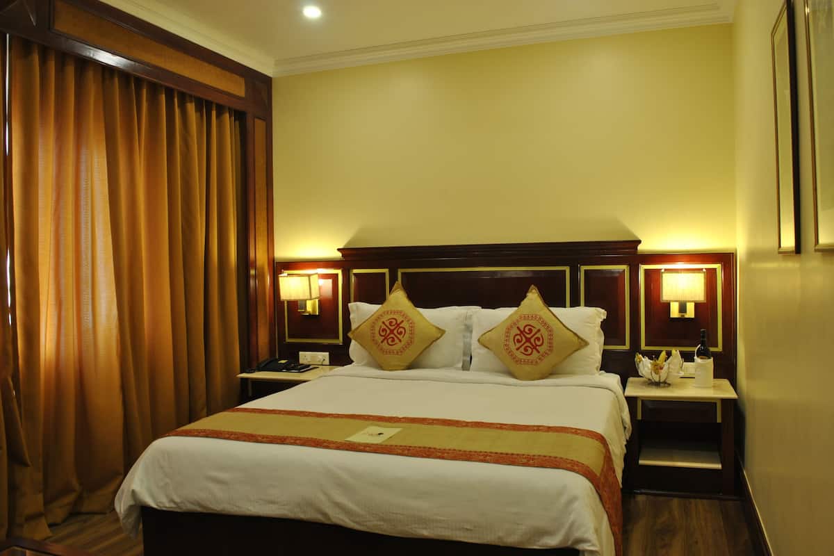 Quarto de hotel com uma cama grande, duas almofadas decorativas, mesa de cabeceira com abajur e cabeceira de madeira.