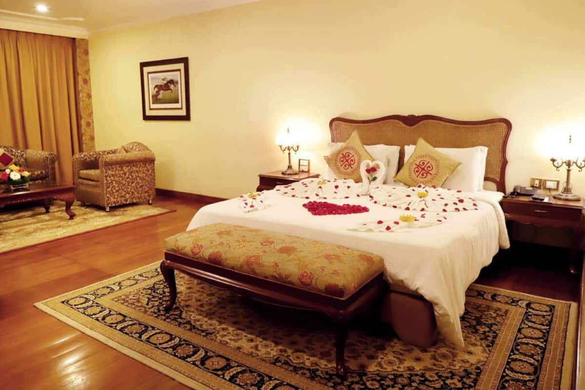 Quarto de hotel com uma cama grande, um espaço de estar com sofá e poltrona, um banco de madeira e um tapete estampado.