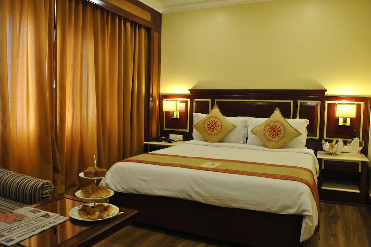 Quarto de hotel com uma cama grande, duas luminárias laterais, uma mesa de cabeceira com abajur, uma mesa de apoio com doces e uma janela com cortinas.