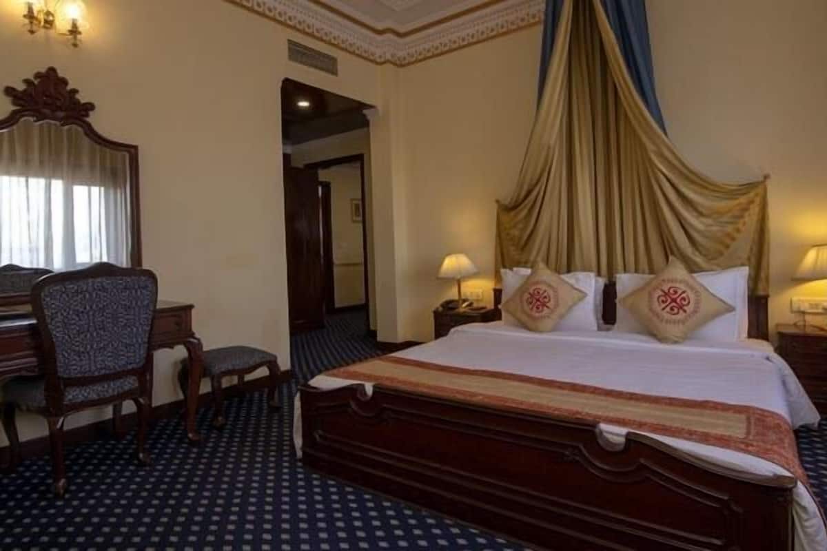 Quarto de hotel com uma cama grande, uma escrivaninha com cadeira, duas luminárias de cabeceira e um dossel.