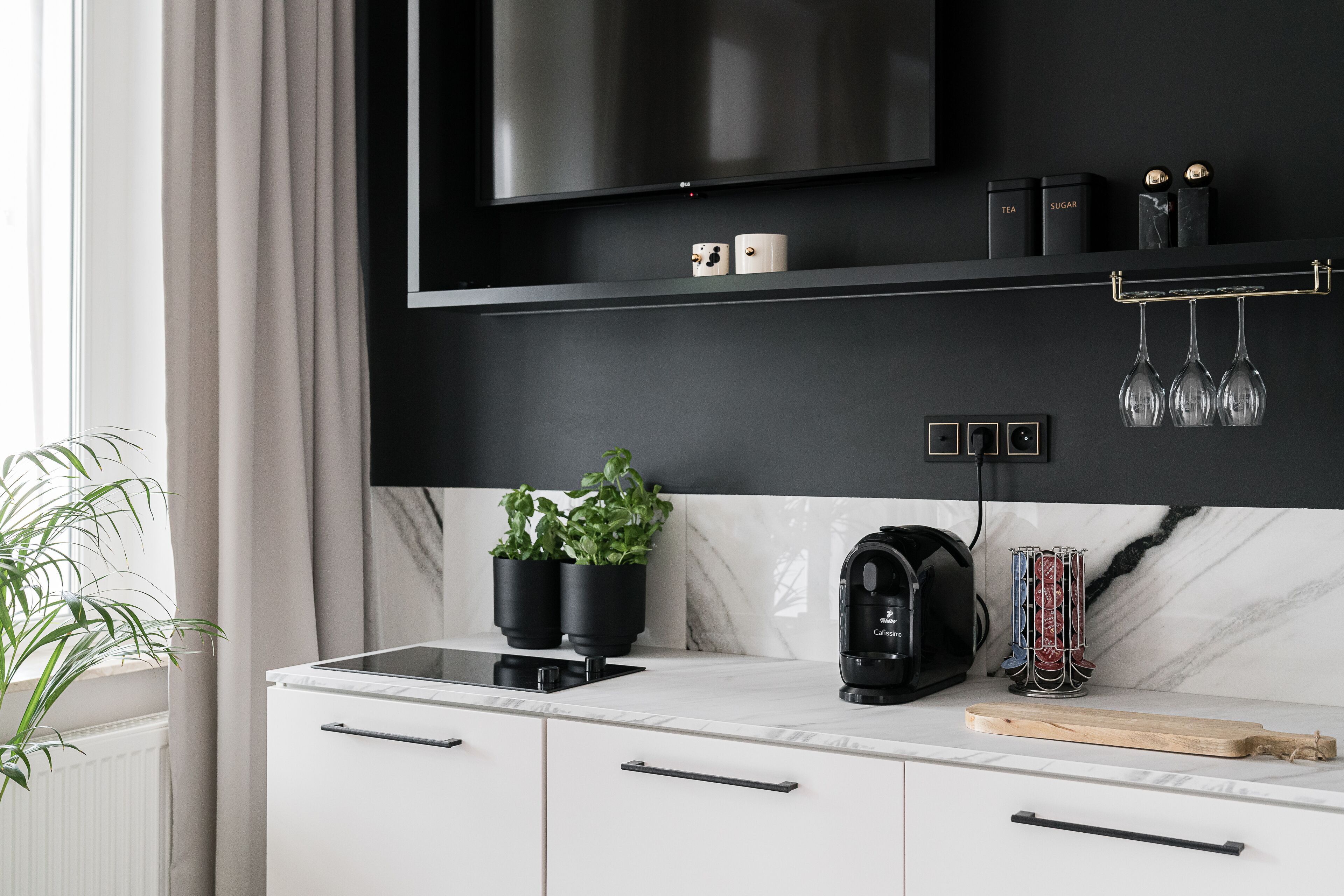 Uma cozinha moderna com uma TV preta fixada acima de uma bancada de mármore, uma cafeteira e uma planta.