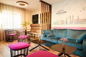Quarto de hotel moderno com um sofá azul, cadeiras rosa, uma mesinha e um mural com vista para a cidade.