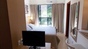 Quarto de hotel com cama, escrivaninha com computador, cadeira, janela com cortinas e ar condicionado.