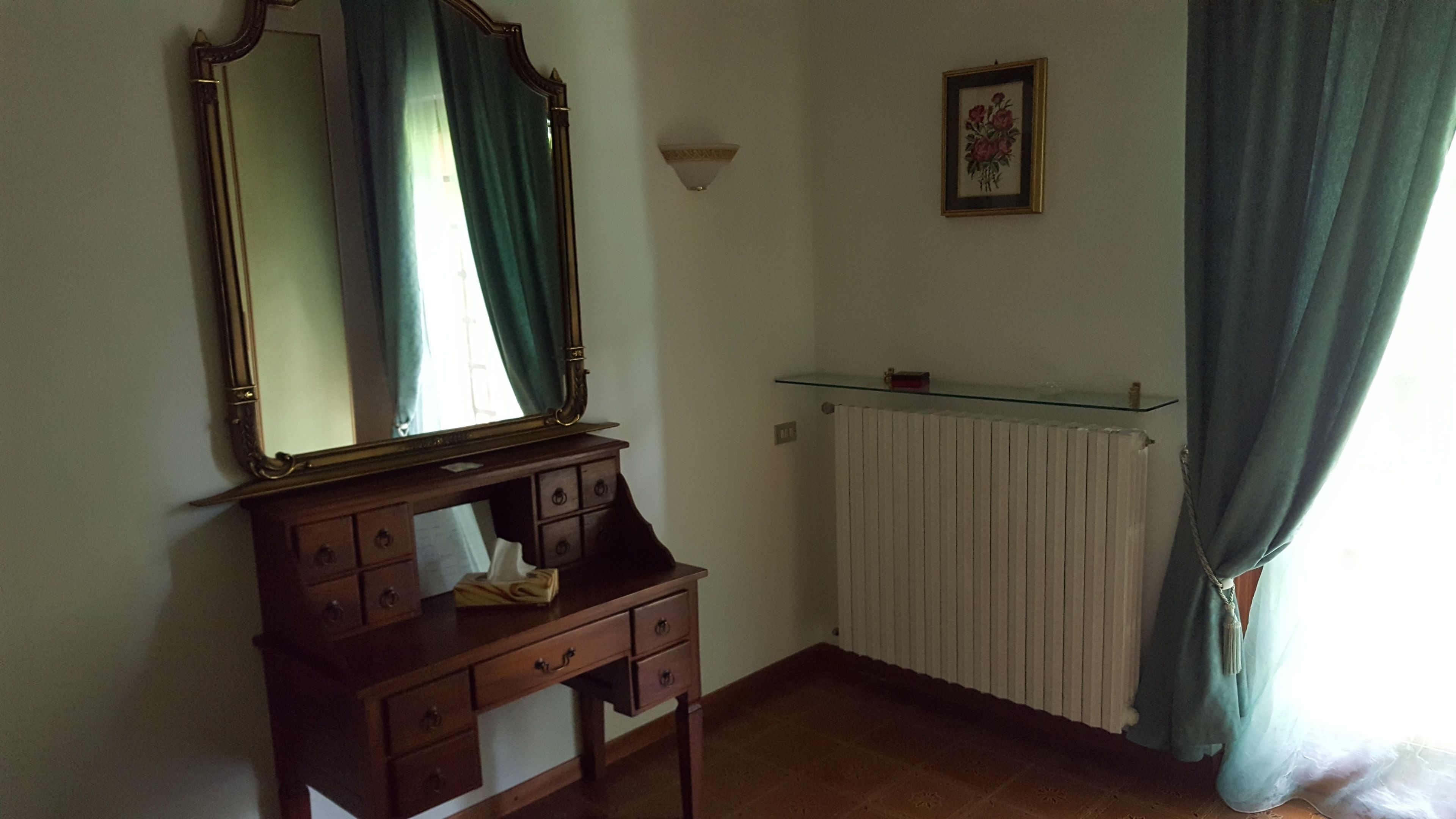 Um quarto com cômoda de madeira, espelho, radiador, prateleira de vidro e quadro na parede.