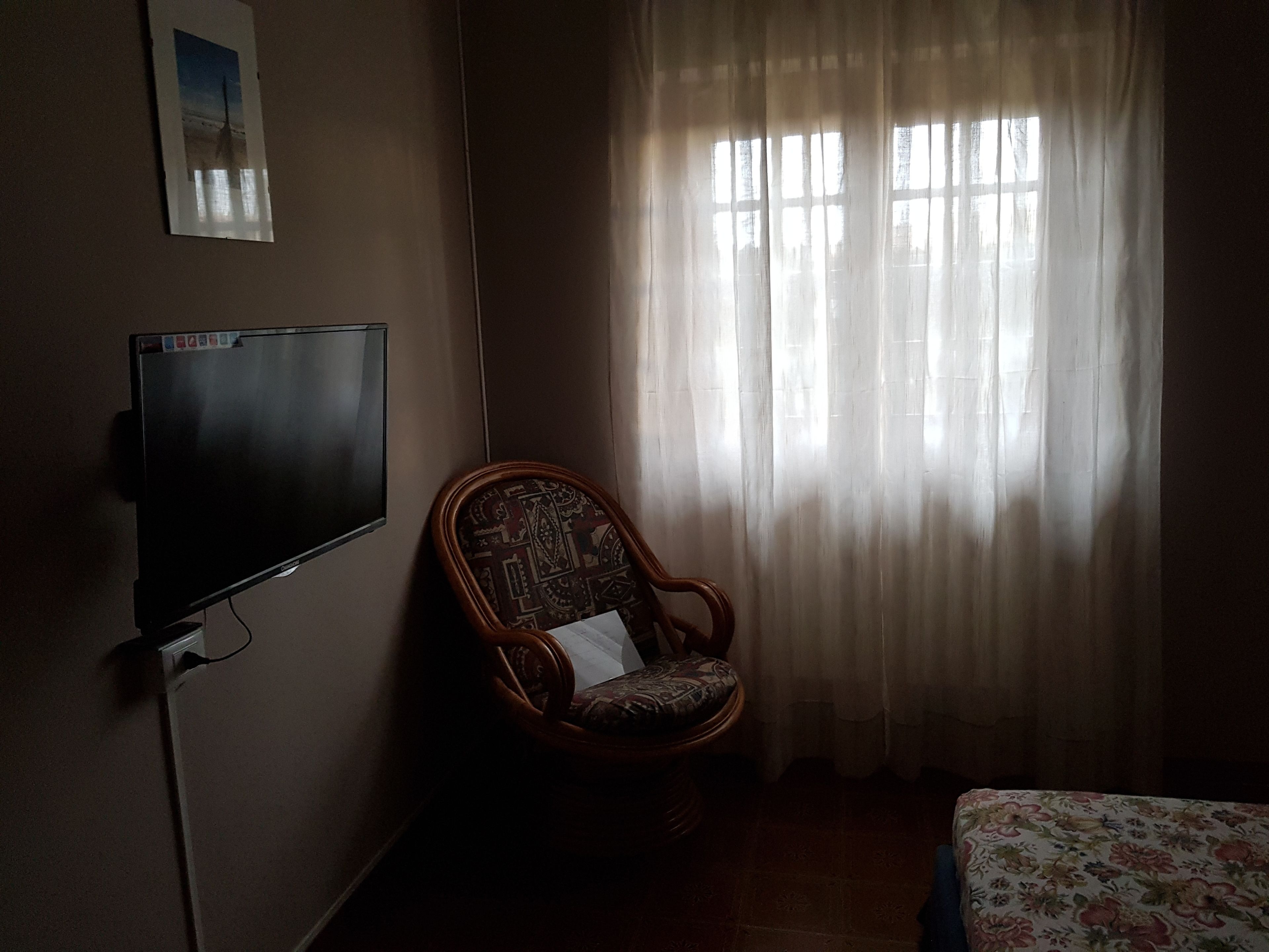 Quarto com televisão na parede, cadeira de madeira com almofada, cama com colcha florida e janela com cortinas leves.