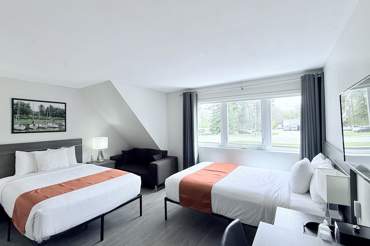 Quarto de hotel com duas camas, uma escrivaninha, uma cadeira, janela com cortinas e um quadro na parede.