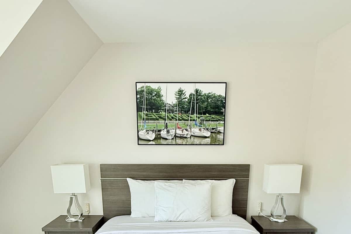 Quarto com cama, duas mesinhas de cabeceira, um criado-mudo, um quadro na parede e teto inclinado.