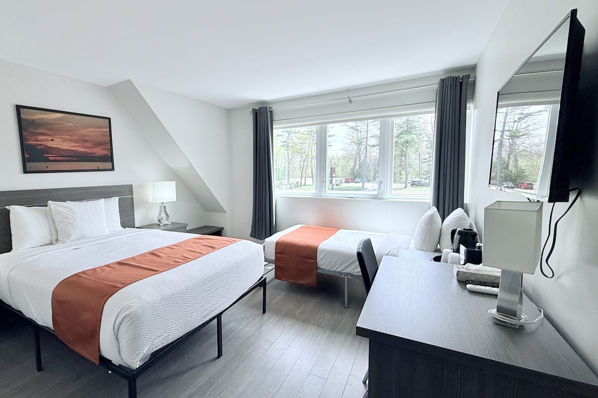 Quarto de hotel com duas camas, uma escrivaninha, uma cadeira, uma televisão e um quadro na parede.