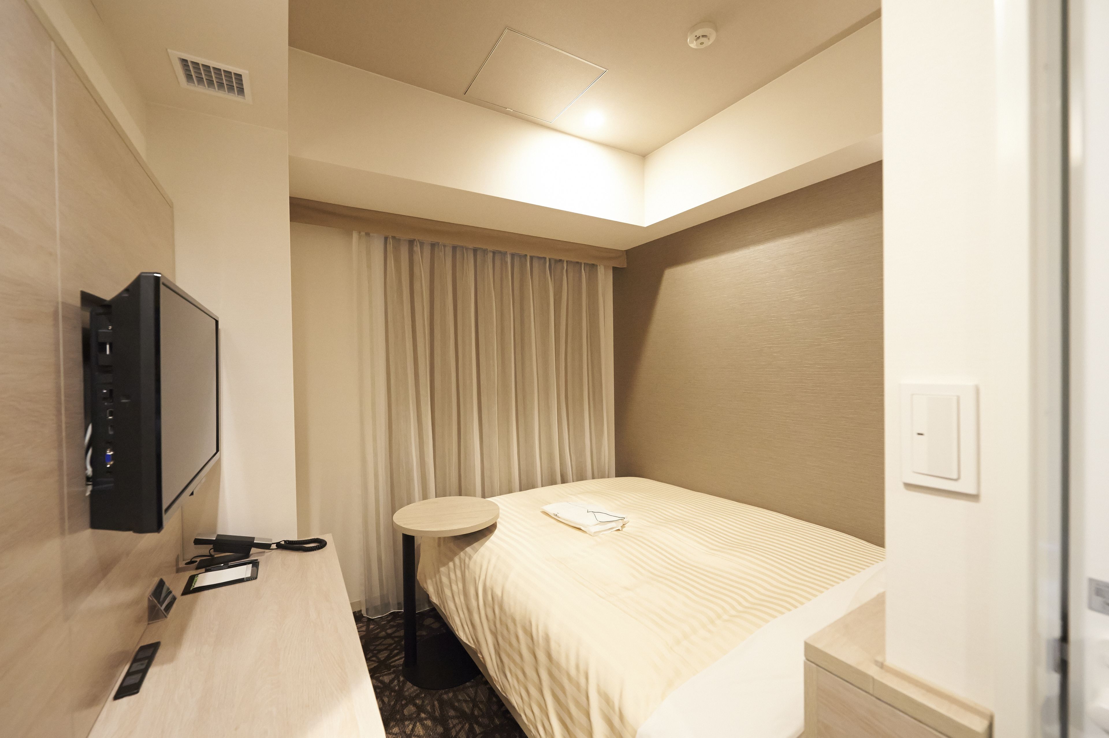 Quarto de hotel com uma cama, uma escrivaninha, uma televisão, cortina e luminária.