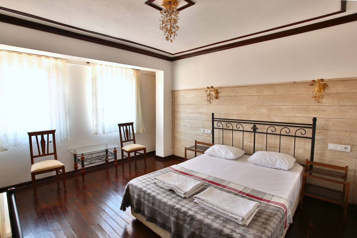 Quarto bem arrumado com uma cama grande, duas cadeiras, uma mesinha e cabeceira de madeira.