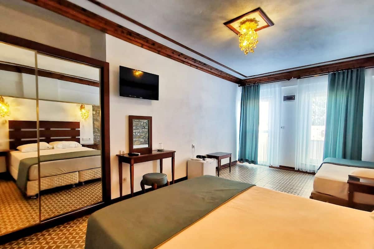 Quarto de hotel com cama, escrivaninha, cadeira, espelho, televisão e uma janela grande com cortinas.