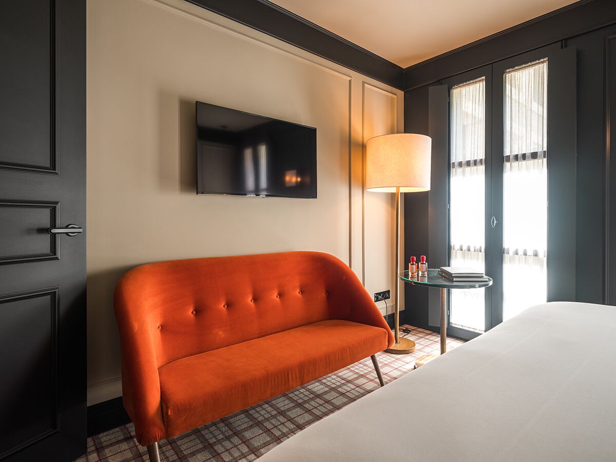 Quarto de hotel com uma cama grande, um sofá laranja, uma TV de tela plana na parede, um abajur e uma mesinha com luminária.