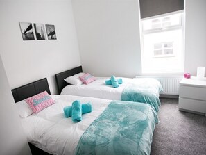 Quarto de hotel com duas camas, cada uma com roupa de cama verde-água e almofadas florais rosa. Há três fotografias a preto e branco na parede, uma janela com persianas e uma escrivaninha branca com um objeto rosa em cima.