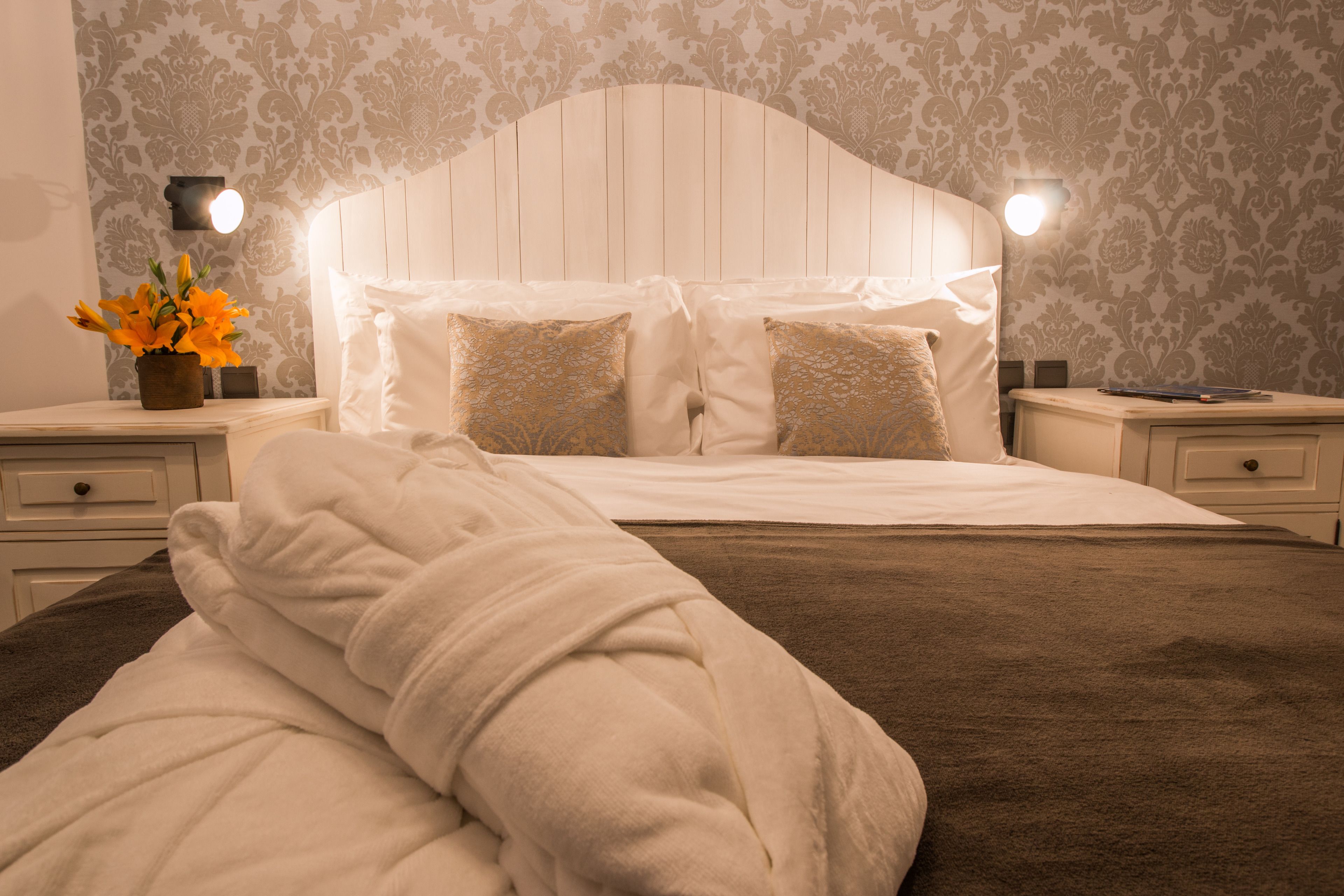 Uma cama bem arrumada com cabeceira branca, duas mesas de cabeceira com abajures e um vaso de flores em uma delas.