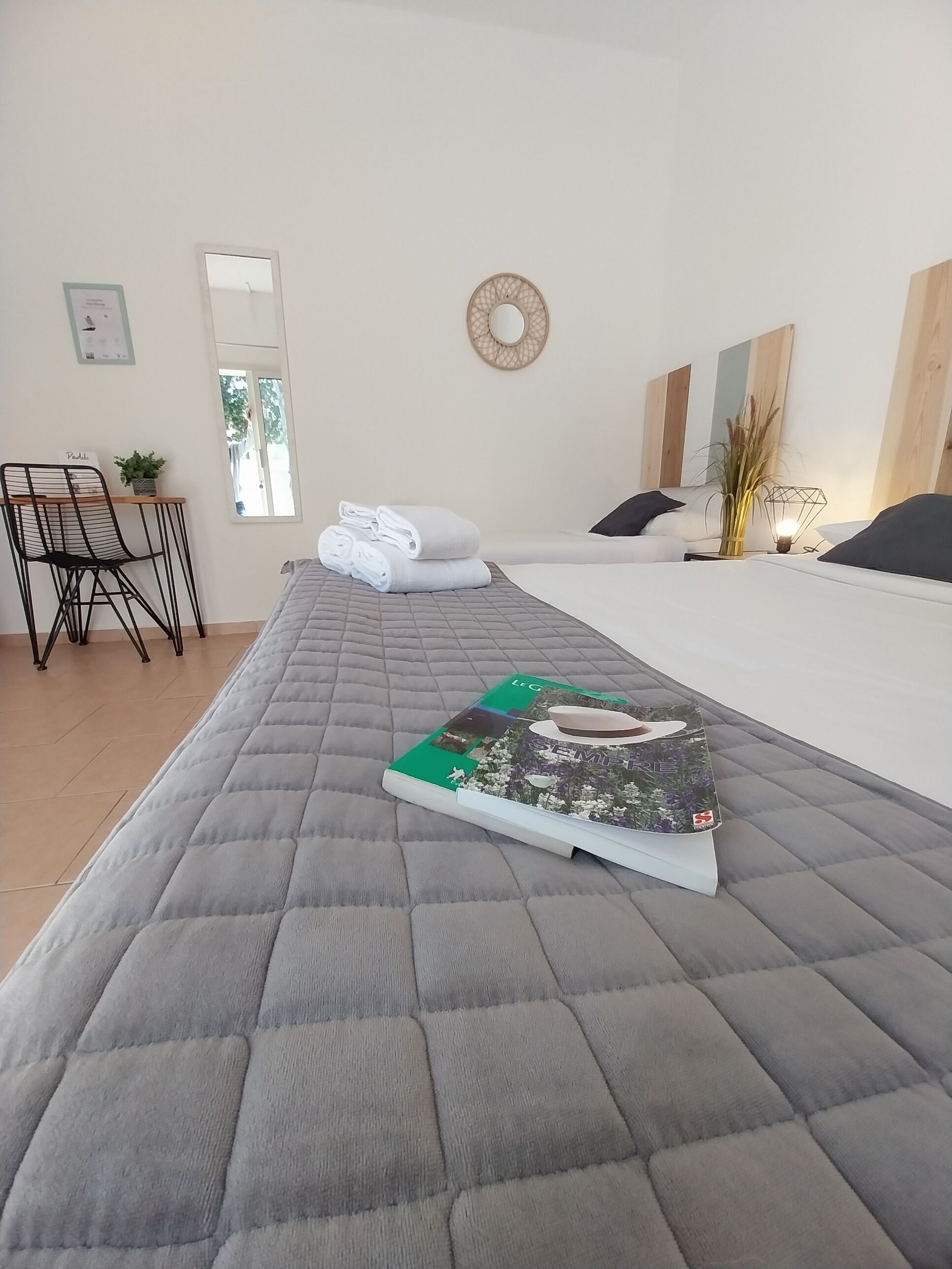 Cama bem arrumada com cobertura cinza acolchoada, uma toalha dobrada e um livro sobre a cama. Há uma pequena mesa com uma cadeira e uma planta em vaso no canto.