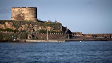 Uma fortificação costeira com uma torre imponente, uma muralha fortificada e um cais que se estende para a água.