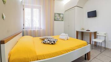 Quarto com cama, escrivaninha, cadeira, televisão e janela com cortinas.