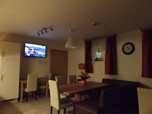 Uma sala de jantar com uma televisão na parede, um relógio e uma janela com cortinas.