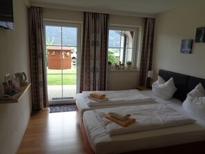 Quarto de hotel com duas camas, uma janela com cortinas, uma mesinha com chaleira e vista para um jardim e um prédio lá fora.