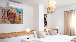 Quarto de hotel com duas camas, cabeceira de madeira, um quadro na parede, um candelabro e uma janela com cortinas.