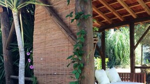 Estrutura de madeira com parede de bambu, cama com almofadas e piso de azulejos.