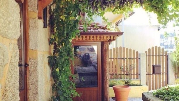 Um edifício rústico de pedra com uma pérgula de madeira coberta de vegetação, uma pequena porta de madeira e um caminho pavimentado que leva a uma cerca de madeira.