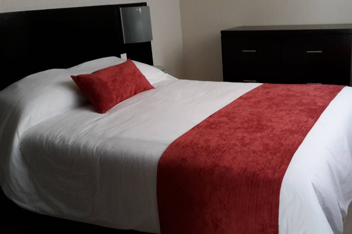Uma cama bem arrumada com colcha branca e vermelha, um travesseiro vermelho e cabeceira preta.