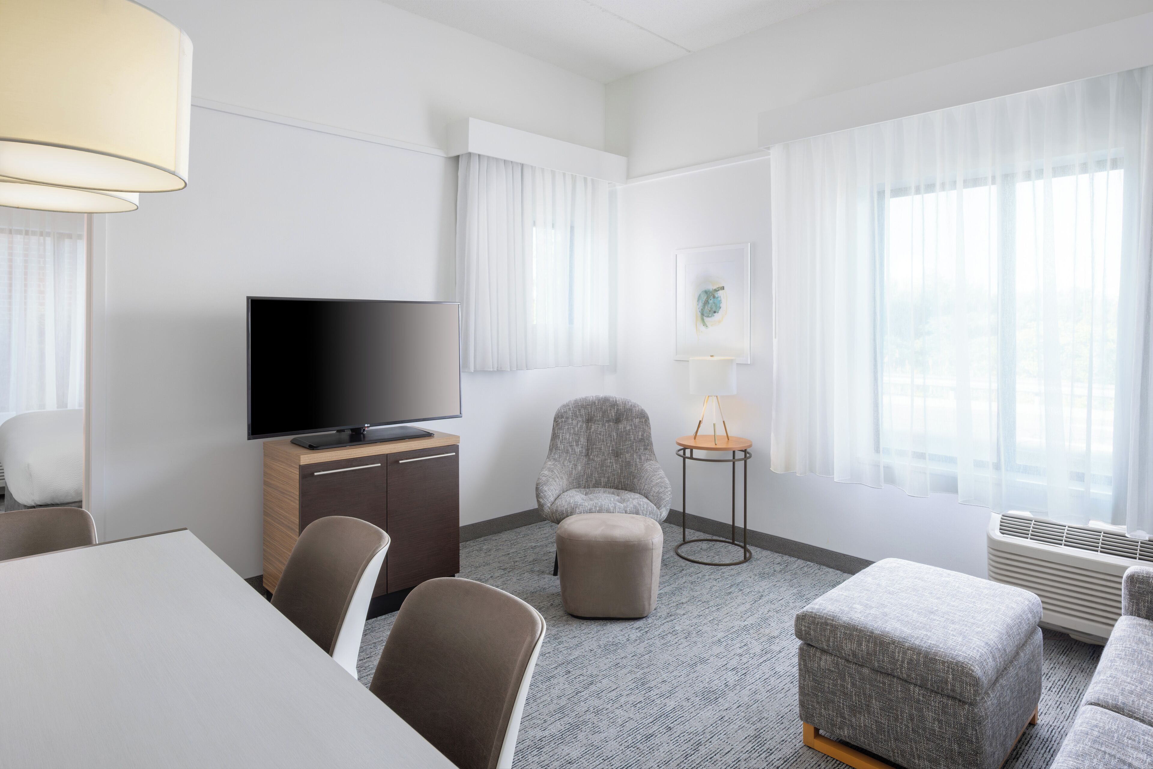 Quarto de hotel com televisão de tela plana, poltrona cinza, mesa lateral redonda com abajur, sofá cinza e mesa de jantar.