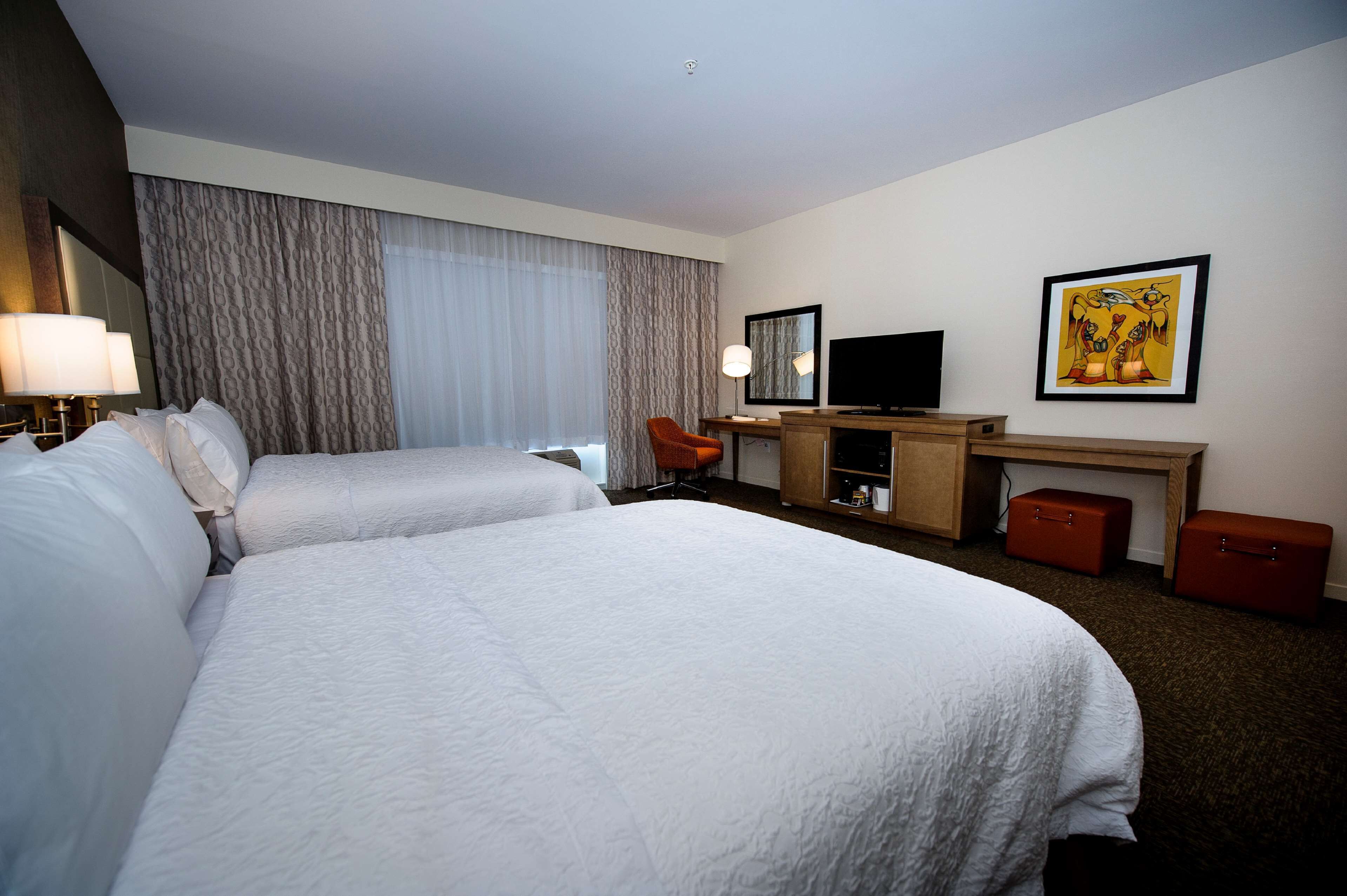 Quarto de hotel com duas camas, uma escrivaninha, uma cadeira, uma televisão e um quadro na parede.