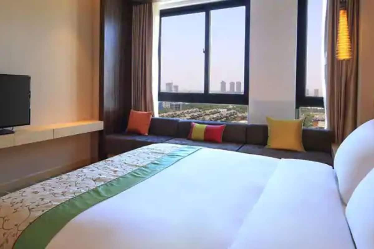 Quarto de hotel com cama grande, televisão de tela plana na parede, janela vista para a cidade e sofá com almofadas coloridas.