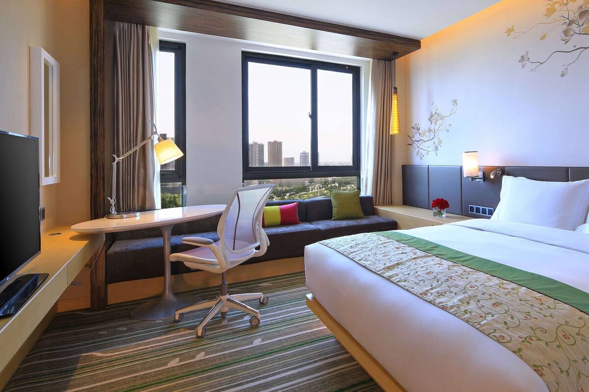 Quarto de hotel com cama, escrivaninha, cadeira, sofá, televisão e janela com cortinas.