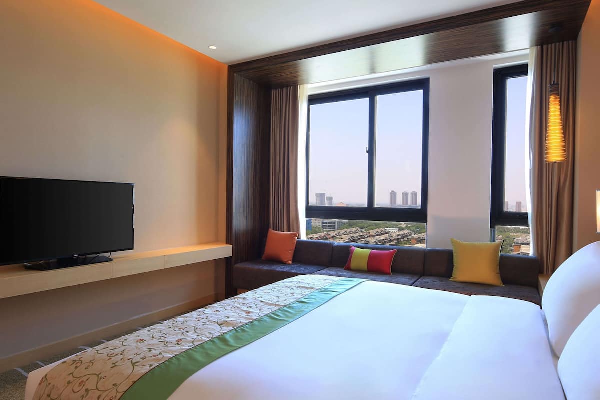 Quarto de hotel com uma cama grande, televisão de tela plana na parede, sofá com almofadas coloridas e vista da cidade por grandes janelas.
