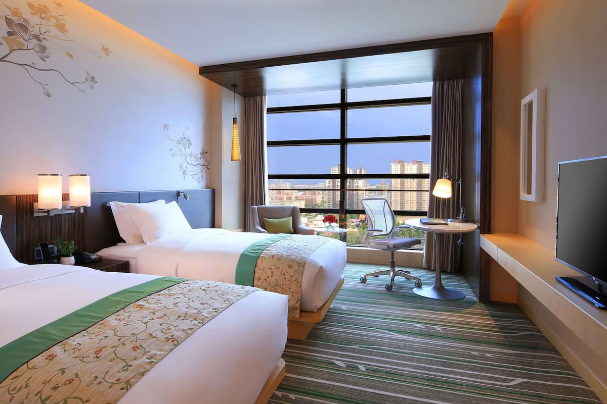 Quarto de hotel com duas camas, uma escrivaninha, uma cadeira, televisão e uma janela grande com vista para a cidade.