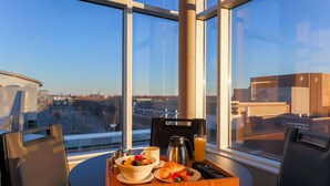 Uma bandeja de café da manhã com croissants, frutas, café e suco sobre uma mesa perto de uma janela com vista para a cidade.
