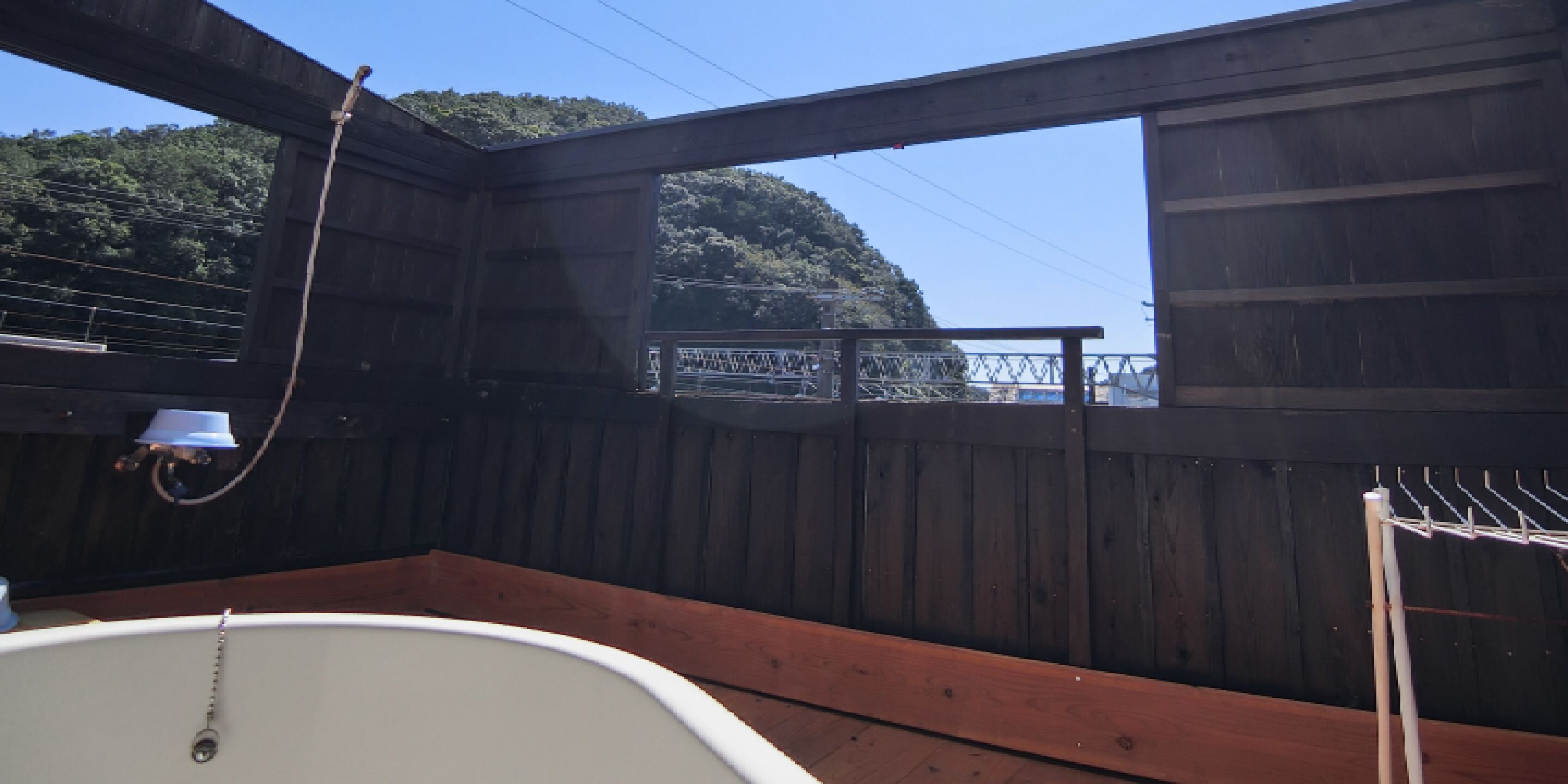 Um deck de madeira com uma banheira branca, guarda-corpo de metal e vista para uma ponte e uma colina arborizada.