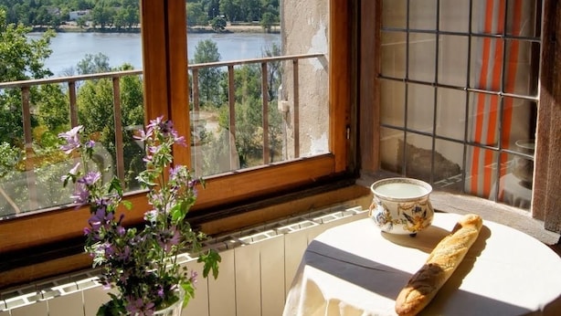 Mesa posta com comida e bebida, um vaso de flores e vista para um rio e horizonte urbano através de uma janela.