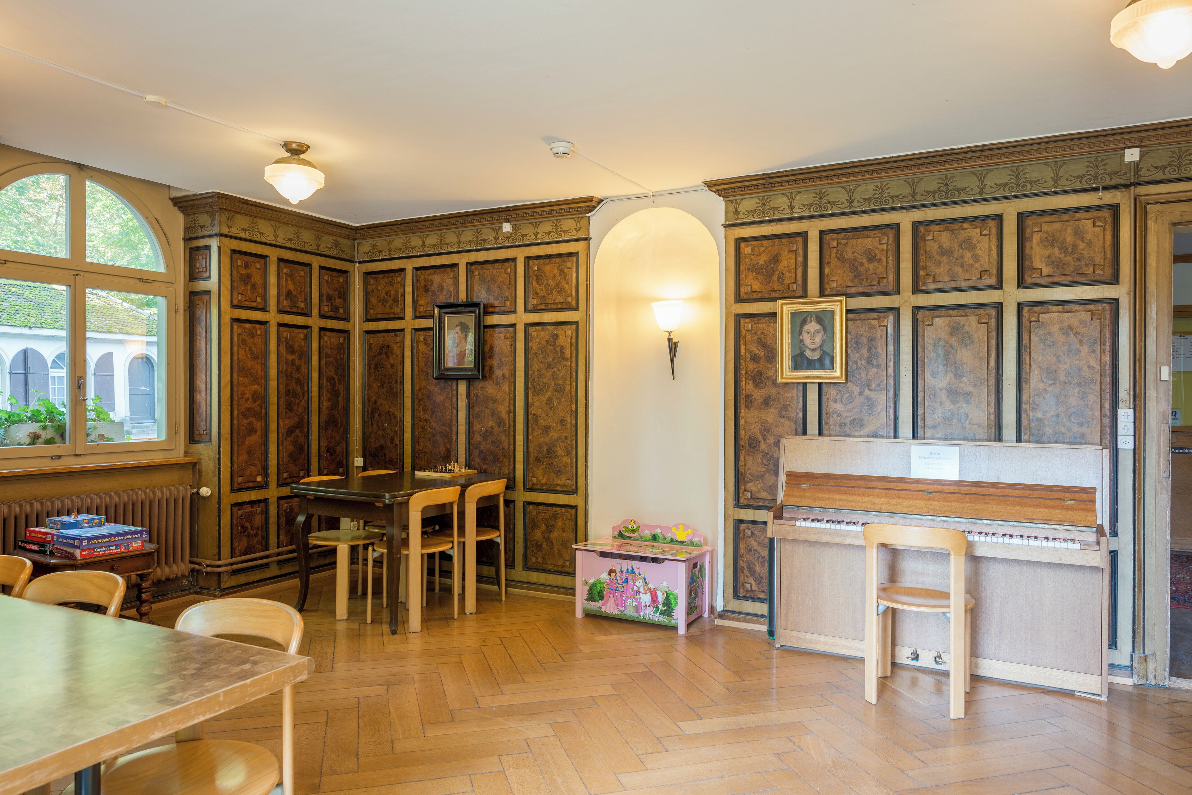 Quarto com piano, móveis de madeira, radiador e janela com vista.