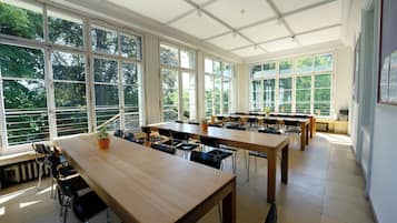 Um quarto amplo com grandes janelas, várias mesas e cadeiras dispostas para refeições ou reuniões.