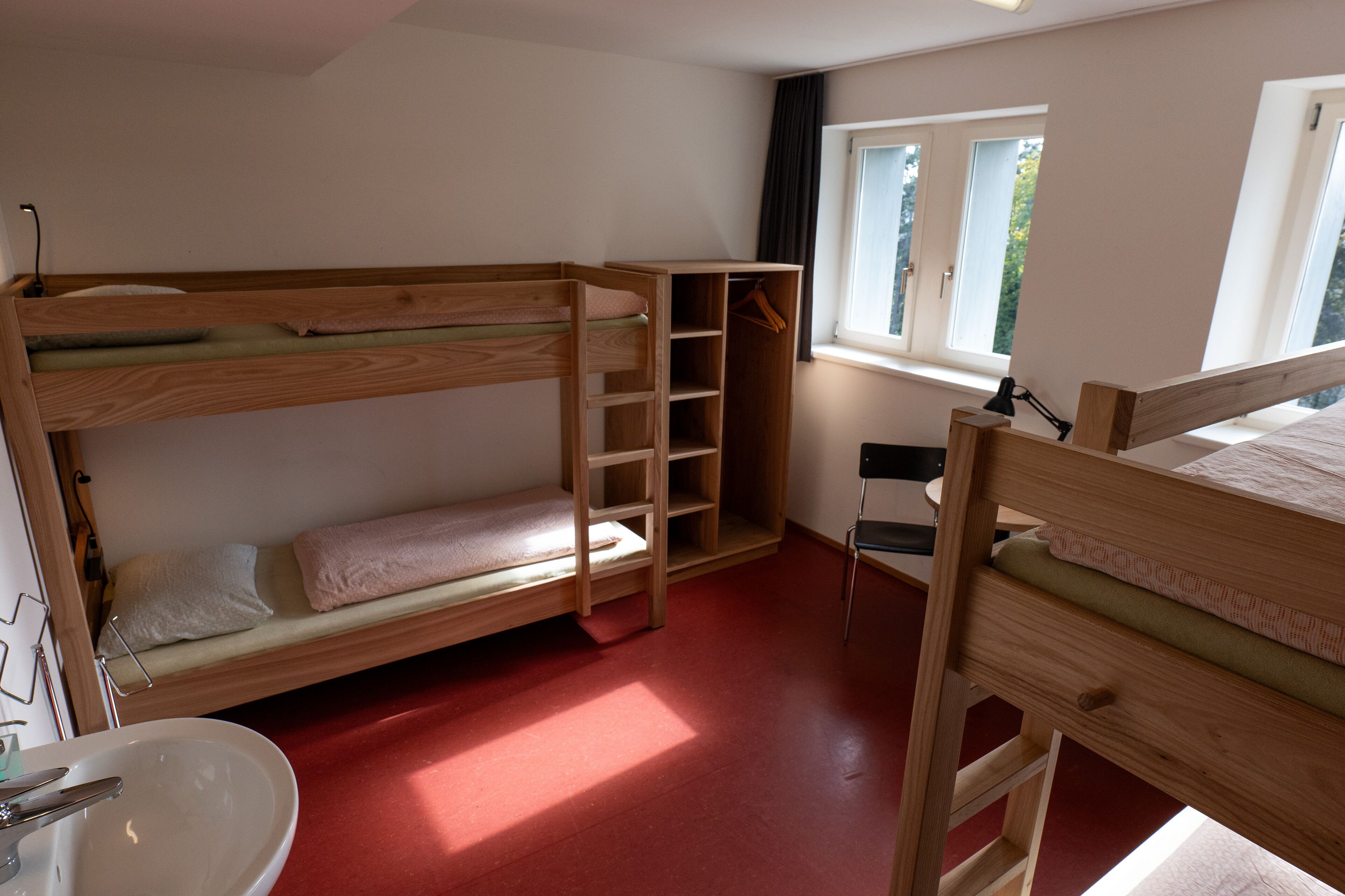 Quarto com beliche de madeira, uma escrivaninha pequena, uma cadeira e uma janela.