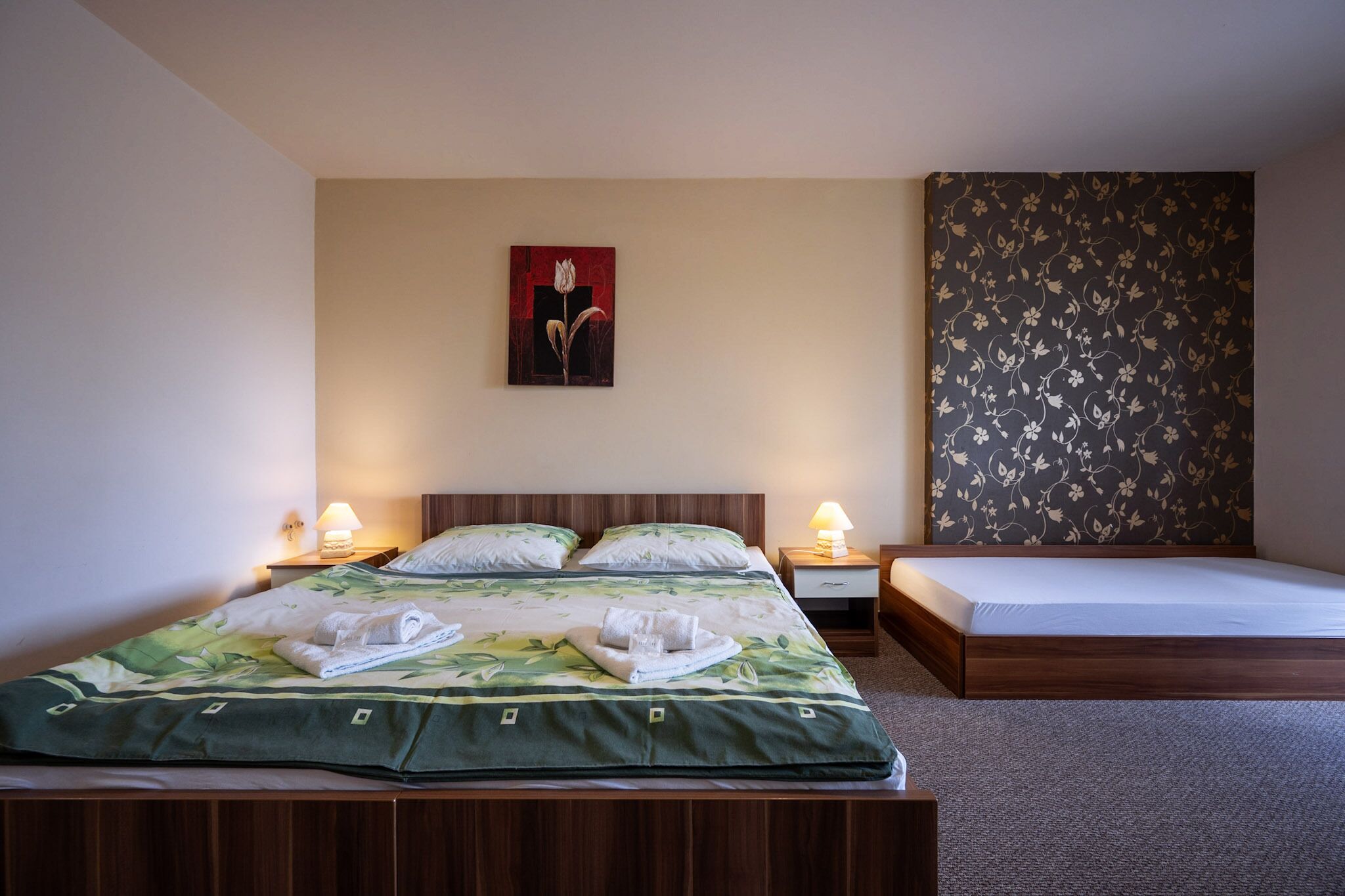 Uma cama bem arrumada com colcha floral verde e branca, duas luminárias de cabeceira, uma mesa de cabeceira, um quadro de flor pendurado na parede e papel de parede com padrão floral na parede adjacente.