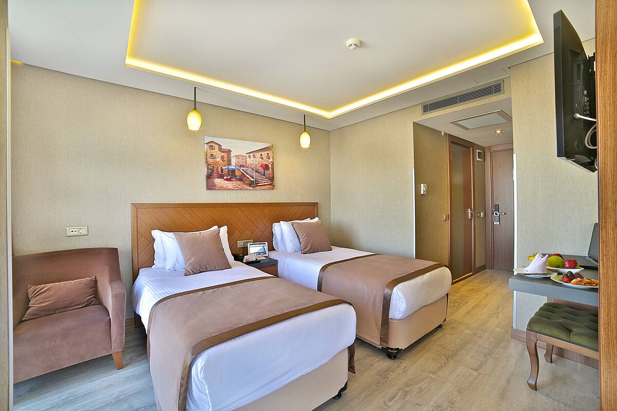 Quarto de hotel com duas camas, uma escrivaninha com um laptop e um espaço de refeições com fruta.