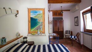 Um quarto com uma cama, uma pintura de uma paisagem costeira, uma prateleira de madeira com um candeeiro e uma janela com vista para o mar.
