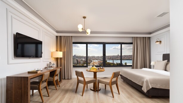 Quarto de hotel com uma cama grande, uma escrivaninha com televisão, uma mesa de jantar com cadeiras e vista para o horizonte urbano por grandes janelas.